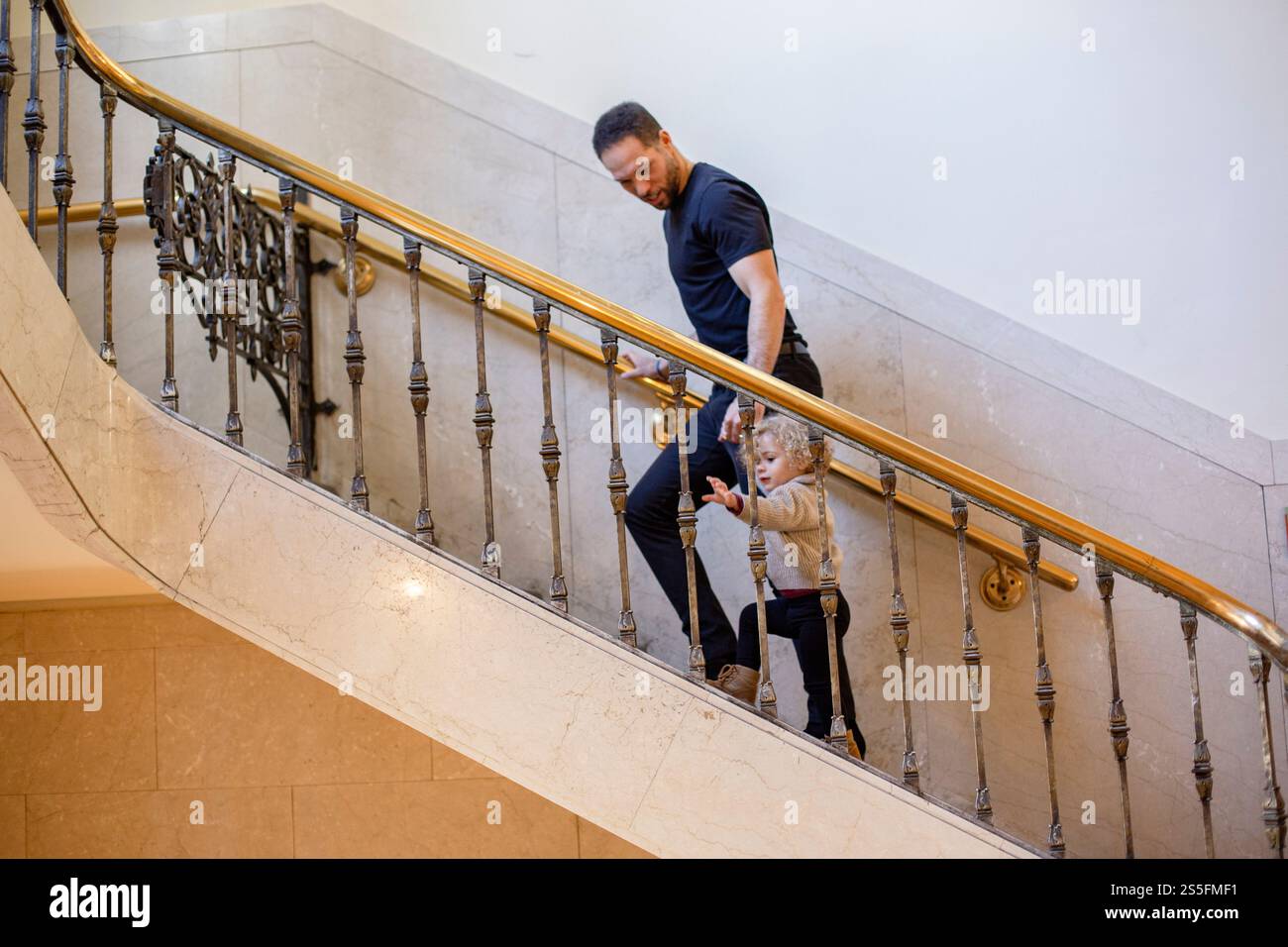 Adult man and young girl holding hands while descending a grand staircase with ornate railing ...