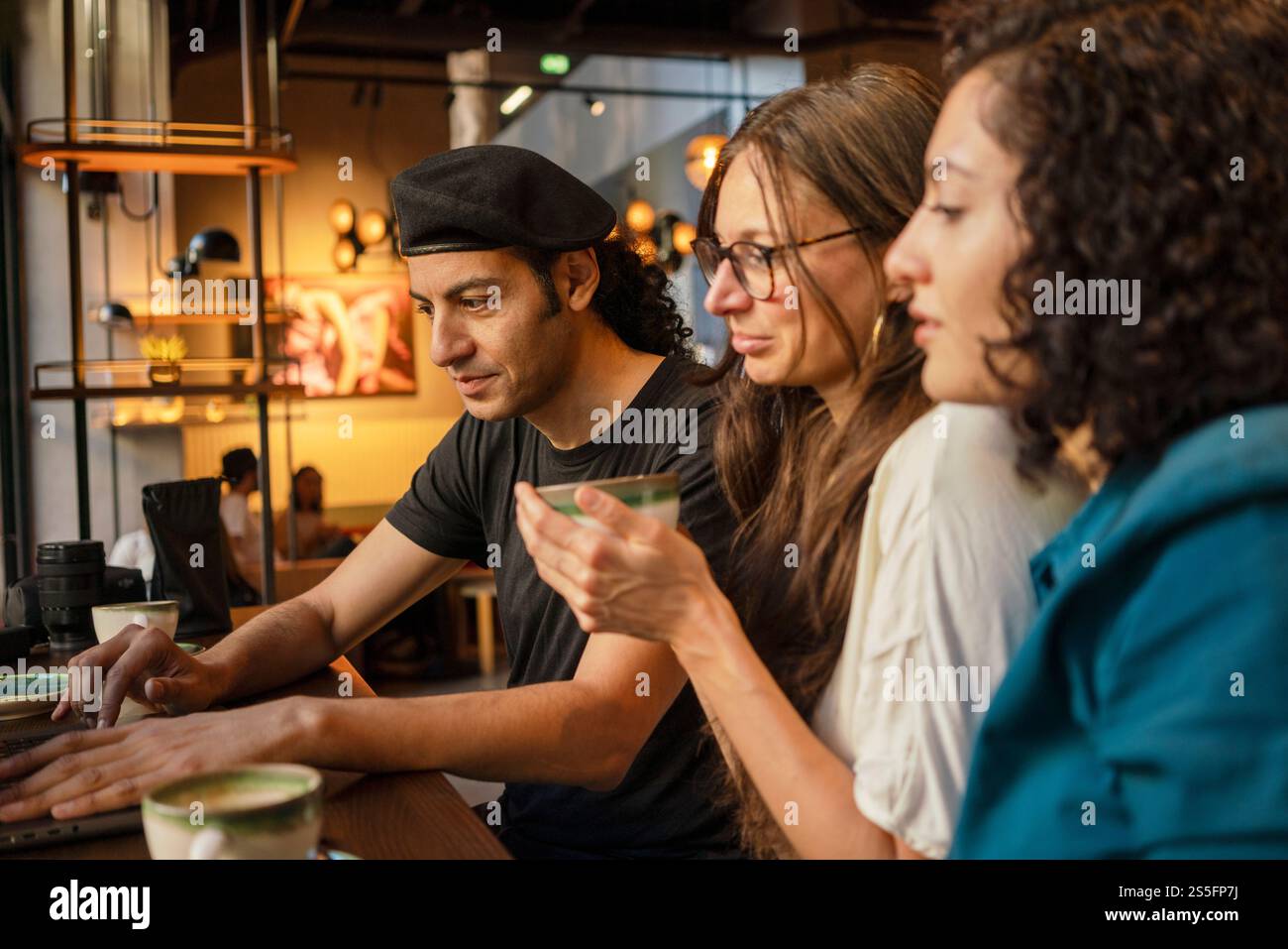 Three adults sit at a cafe table, one man working on a laptop and two ...