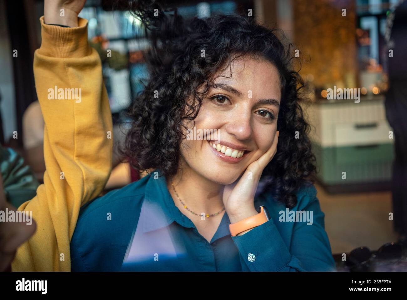 Smiling woman with curly hair resting her chin on her hand by a window ...