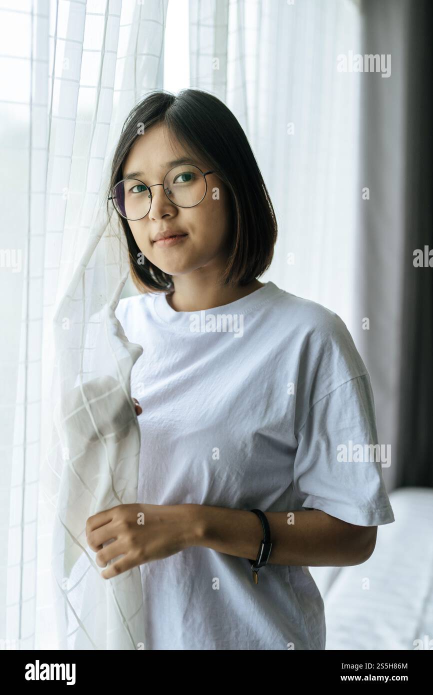 Woman wearing a white shirt, standing, catch a curtain near the window ...