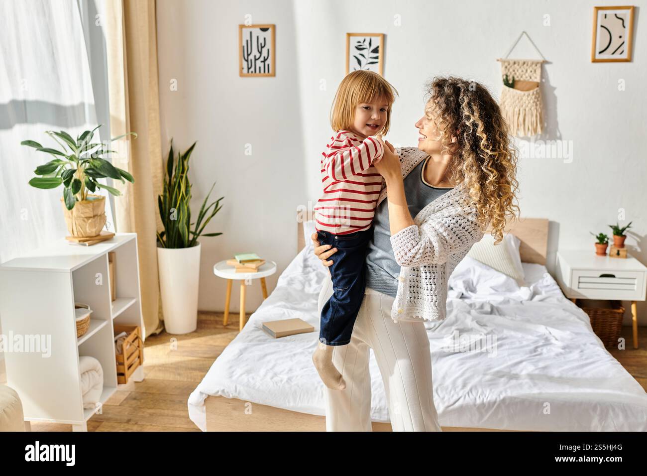 A curly mother joyfully lifts her daughter in a light-filled, stylish ...