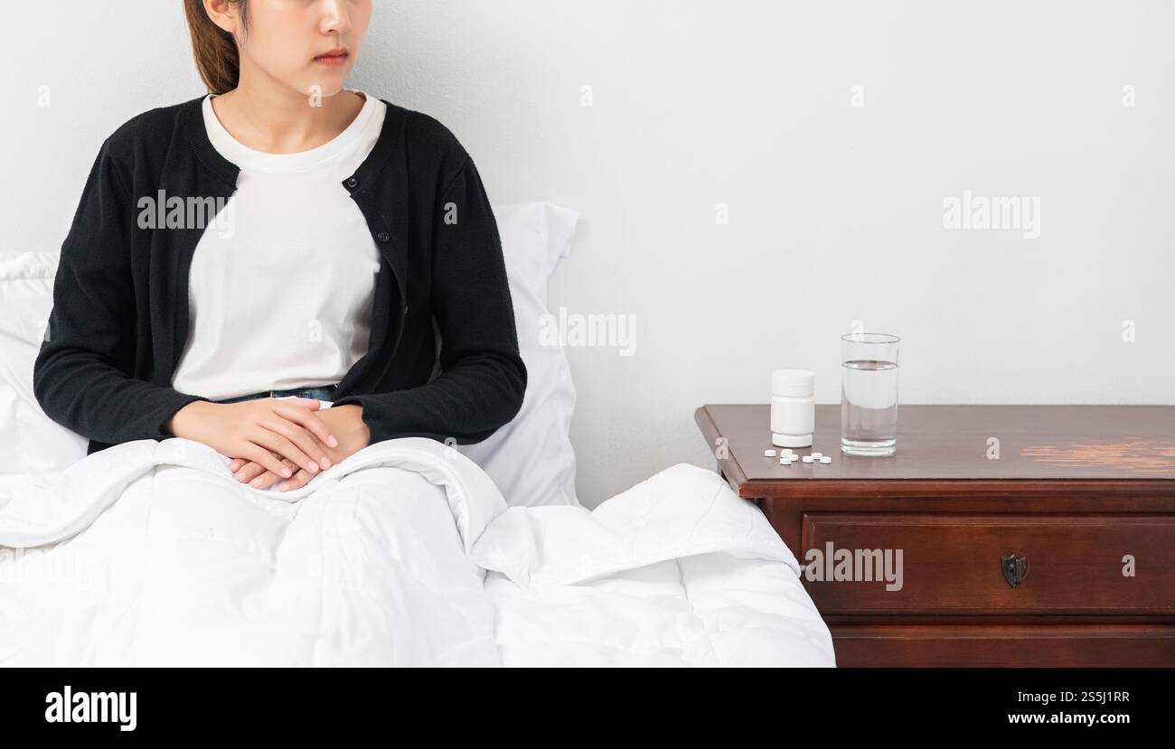 An uncomfortable woman sits on the bed and has medicine on the table ...