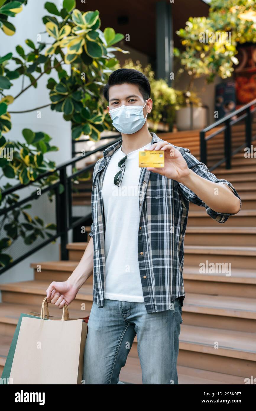 Portrait smiley Young handsome man in mask holding paper bag and show credit card on stair of ...