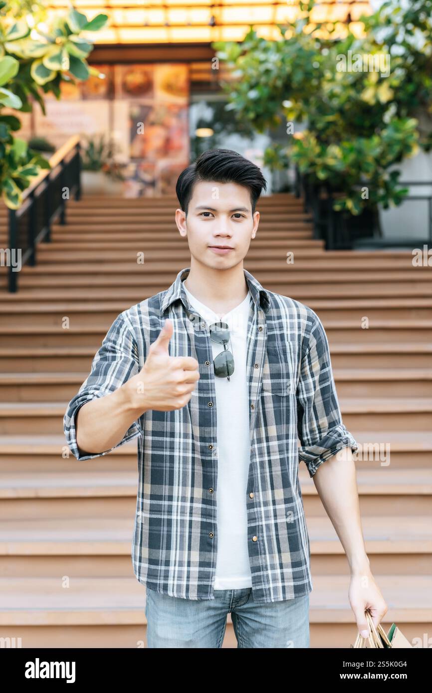 Handsome young man holds multiple paper bag and shows his hand sign thump up. At the stairs of a ...