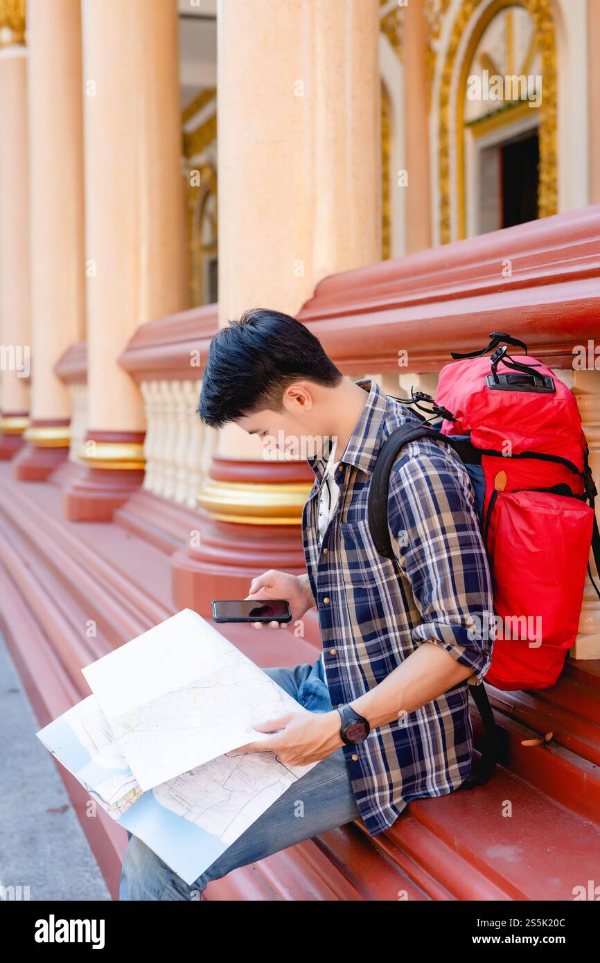 Young Asian backpacker male sitting and hold a paper map and use ...