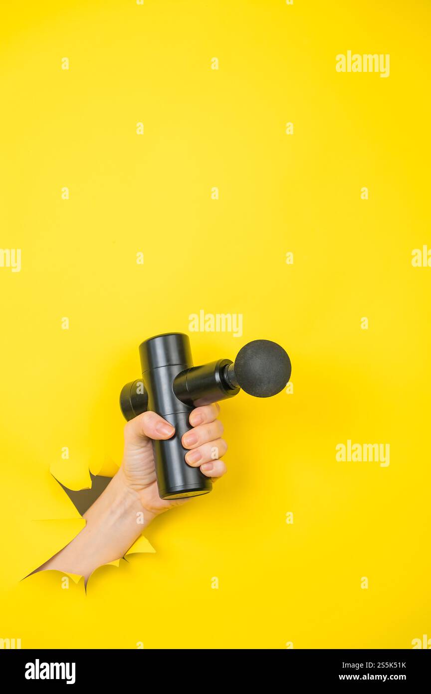 Female hand sticking out with massage gun from yellow paper background ...