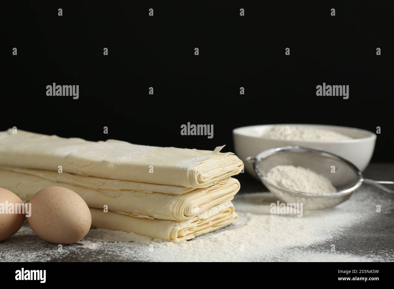Stack of raw puff pastry dough, eggs and flour on dark table against ...