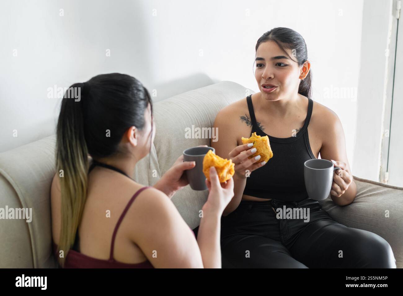 two young latina friends talking while eating their snack, sitting on ...