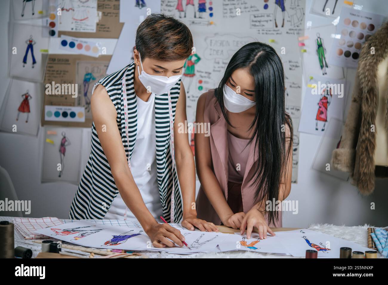Young asian female designer and colleague wearing medical mask during working in modern tailor ...