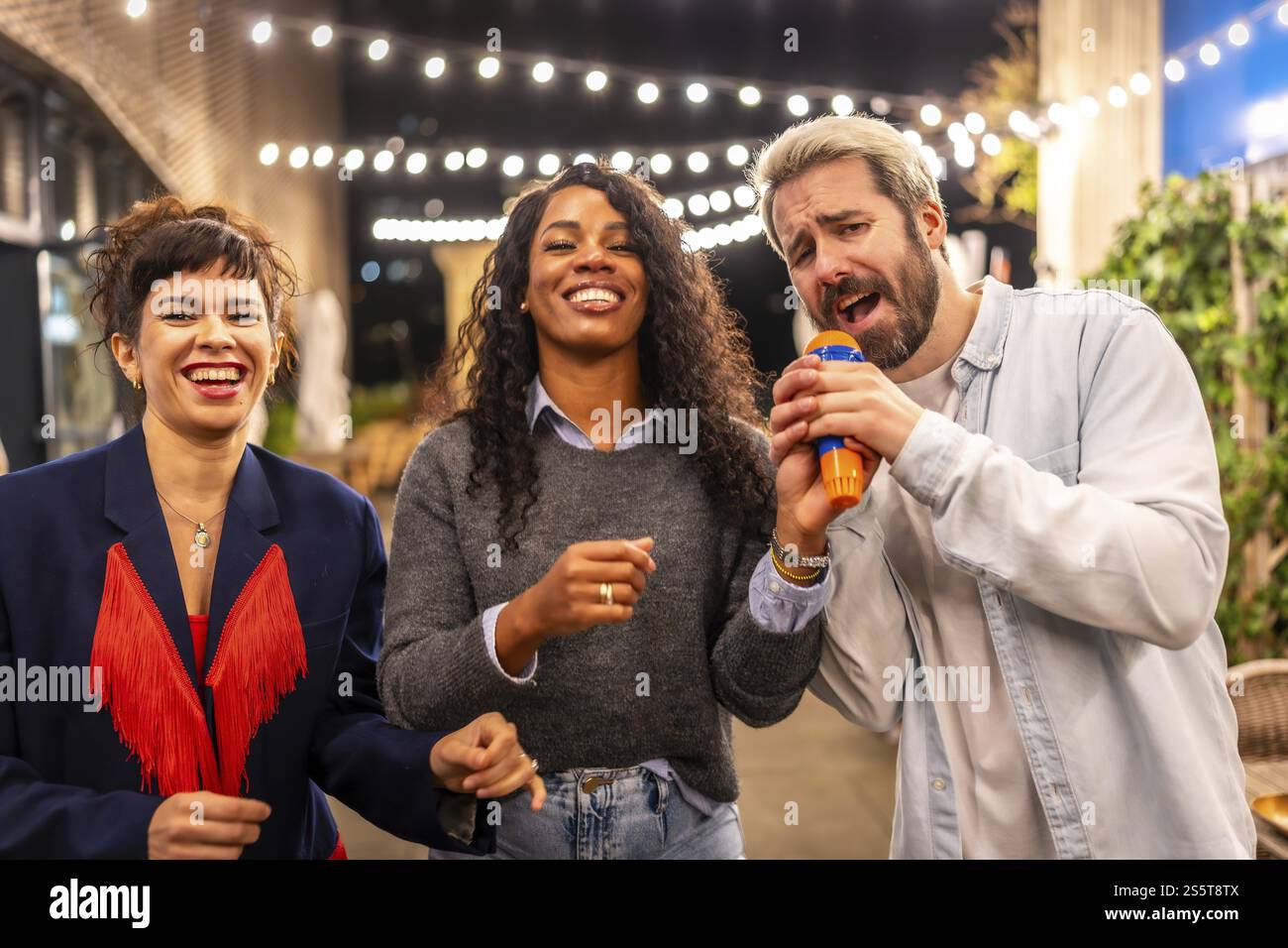 Three diverse friends singing karaoke and laughing at night party in ...