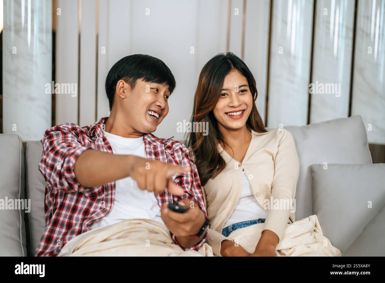 Young Asian couple watch television on sofa in new house and watch funny movie together. They ...