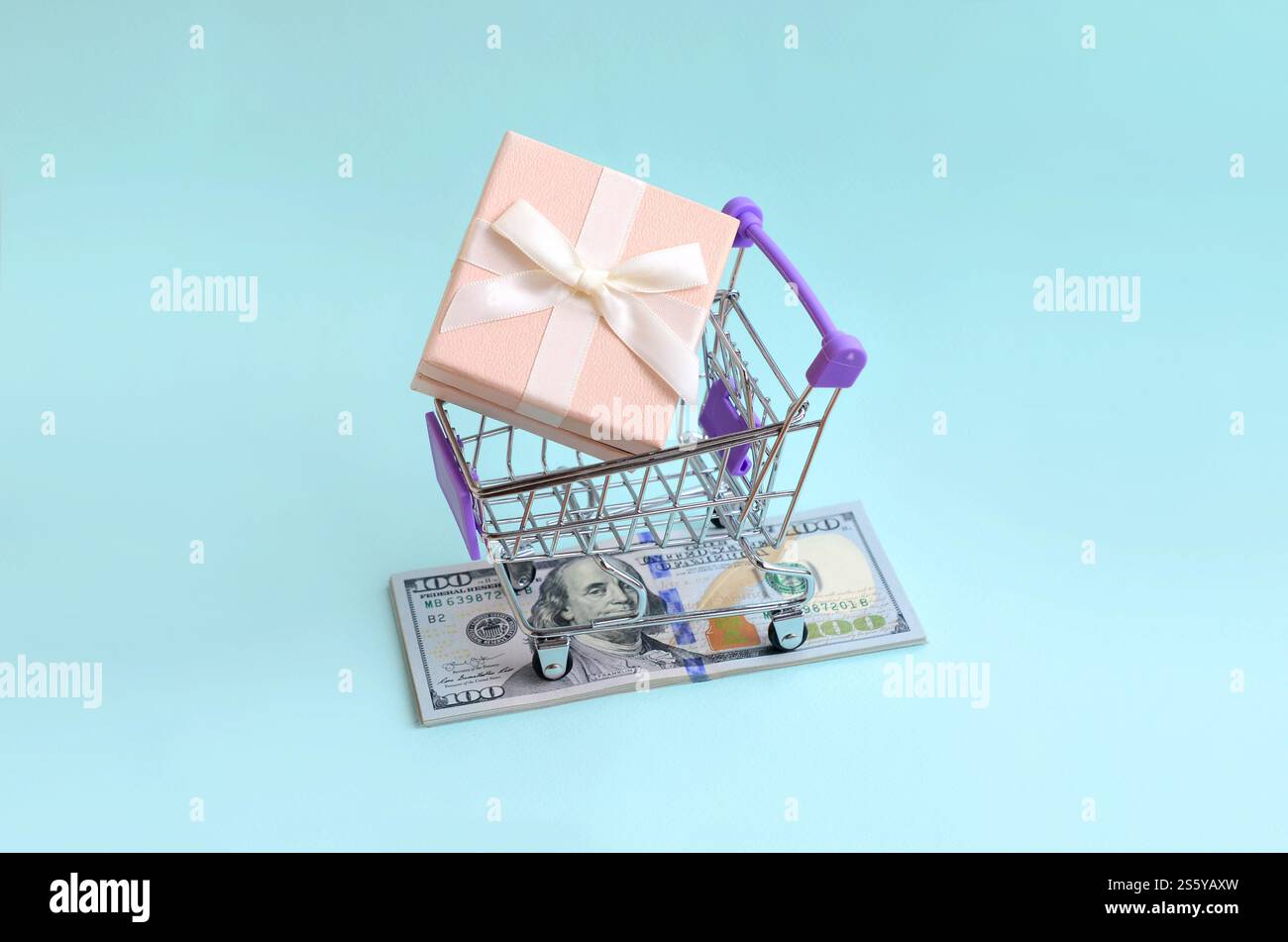 Gift box in a small shopping cart lies on a dollar bills on light blue ...