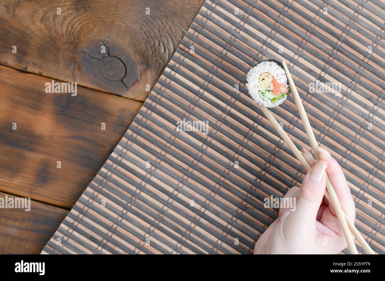 A hand with chopsticks holds a sushi roll on a bamboo straw serwing mat background. Traditional ...
