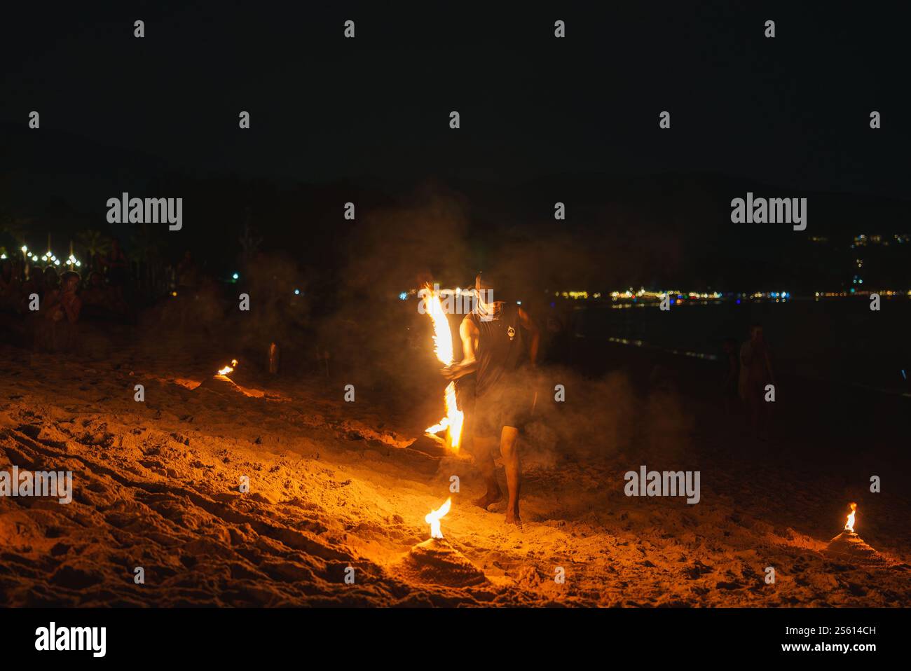 A fire performer manipulates flaming torches on a sandy beach at night ...
