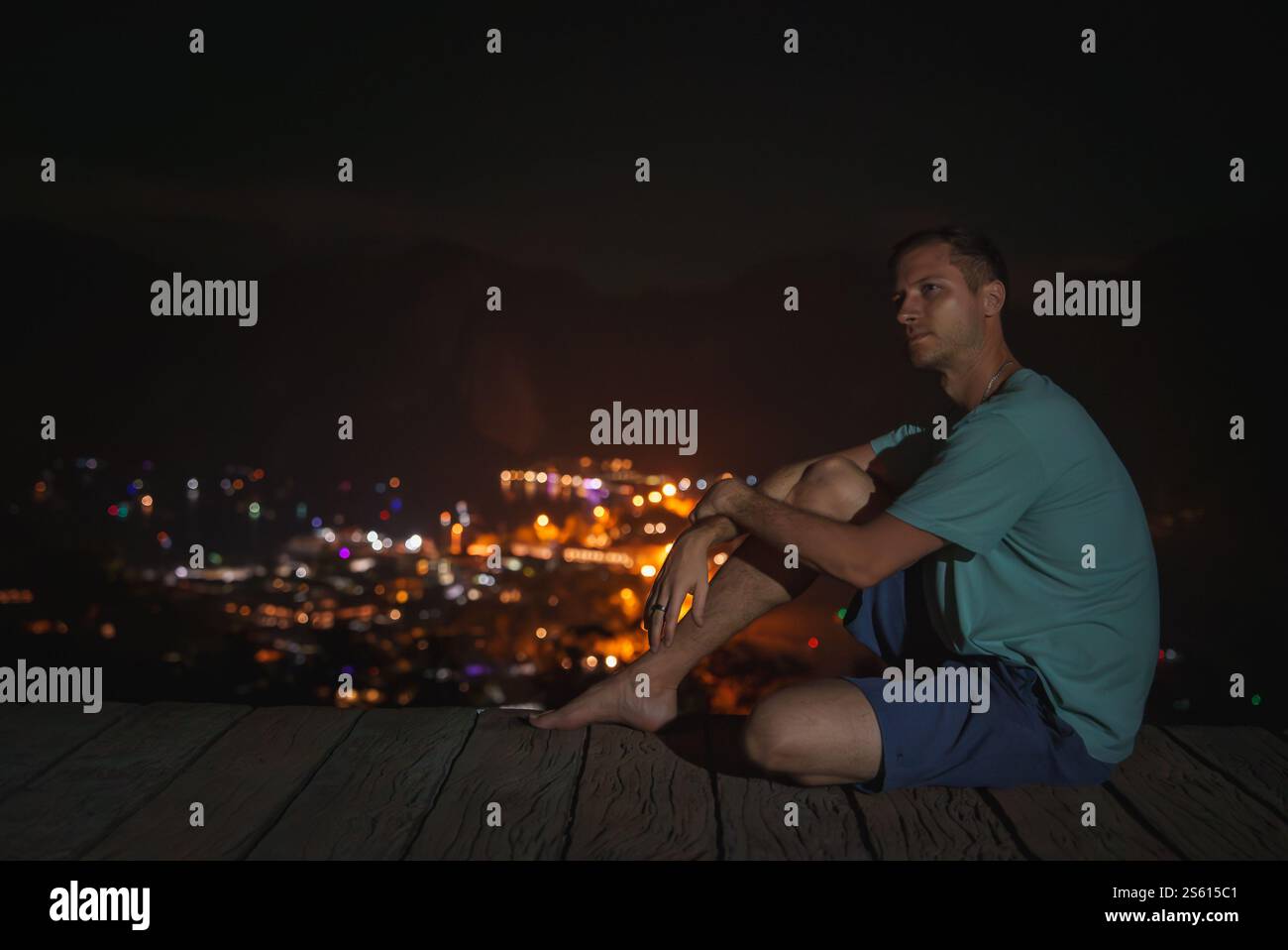 Man Seated on Wooden Platform Overlooking Lit Coastal Town at Night ...