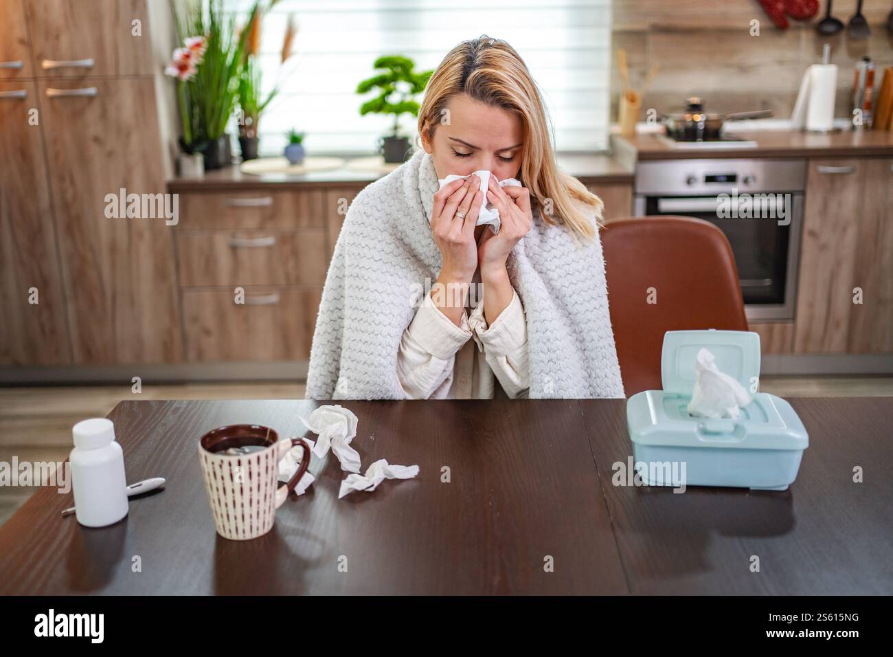 Woman wrapped in a blanket looks unwell at kitchen table during winter ...
