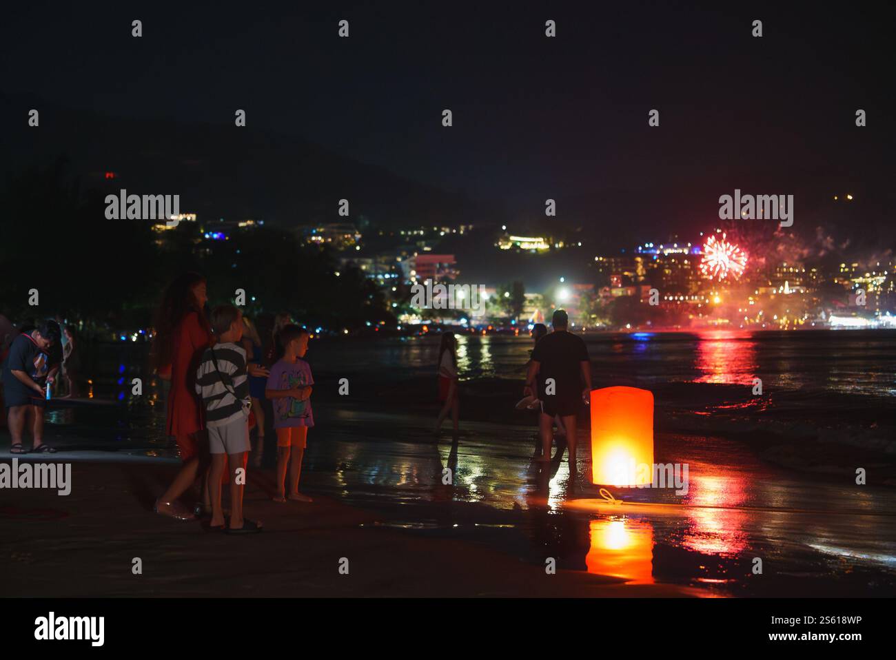 A glowing orange lantern reflects on wet sand as colorful fireworks ...
