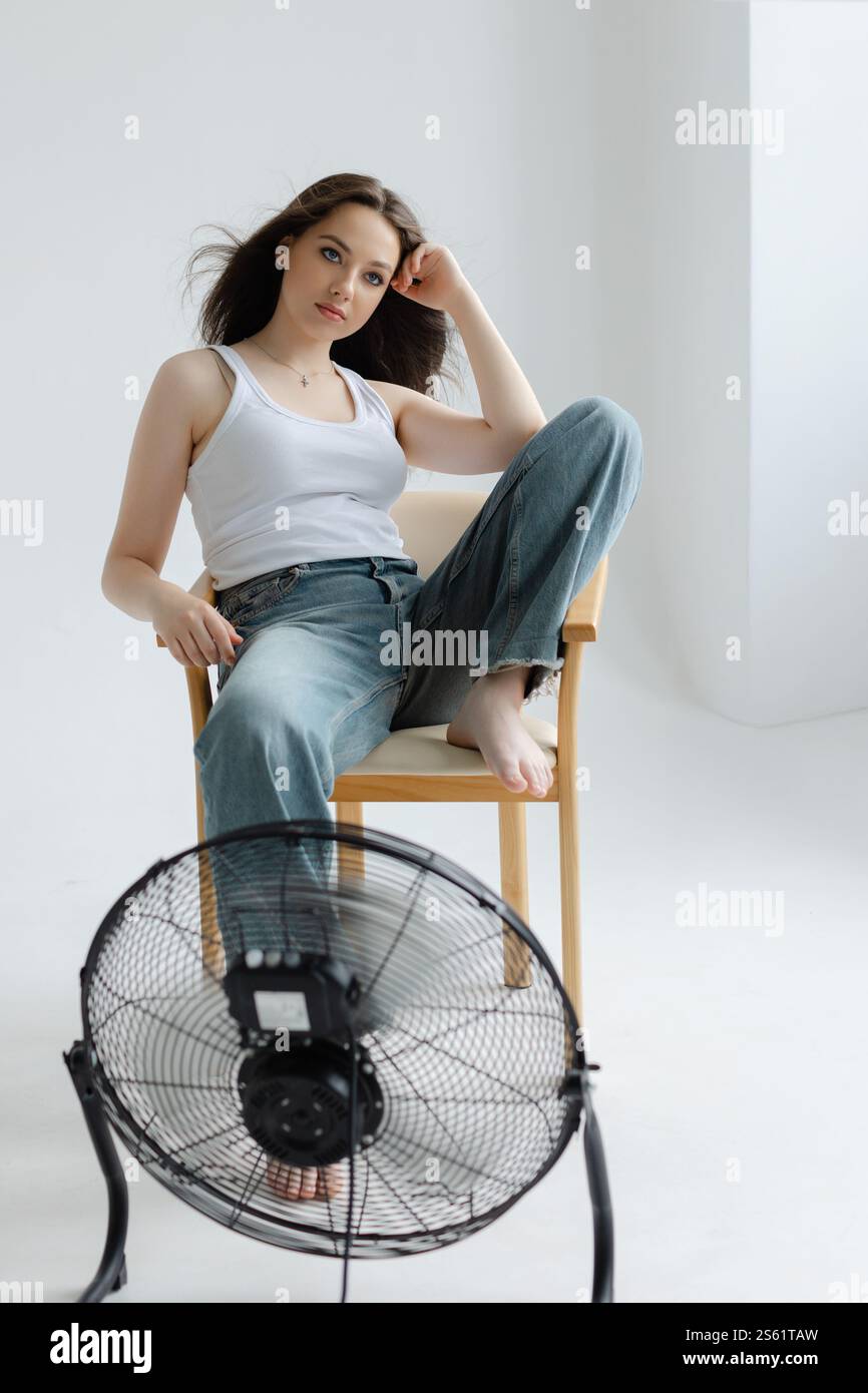 Young woman sitting comfortably in a chair, using a fan to cool off and ...
