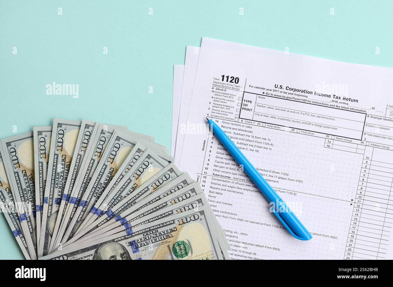 1120 tax form lies near hundred dollar bills and blue pen on a light ...