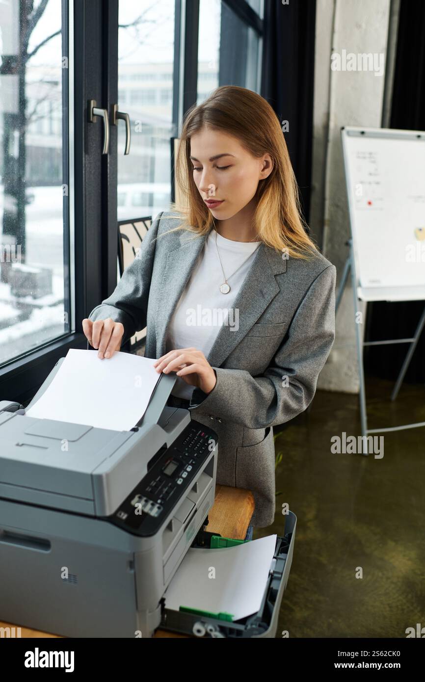 Stylish young woman uses a printer in an office with big windows and ...