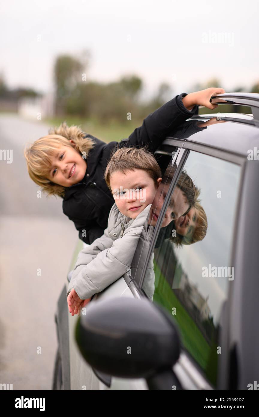 Boys lean out of a moving car’s windows, laughing and enjoying the chilly wind in a small road ...