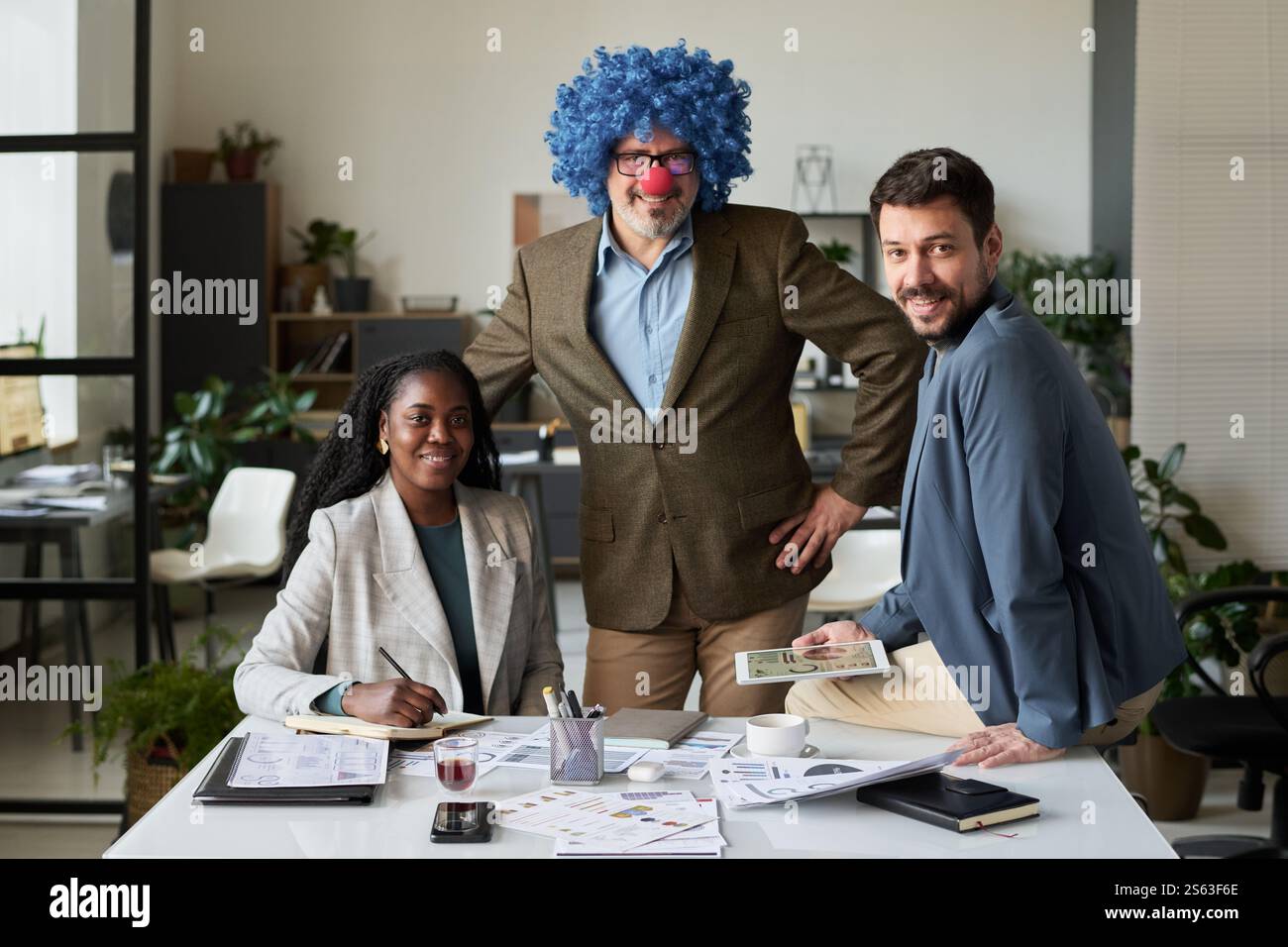 Two happy young intercultural subordinates surrounding mature clown ...