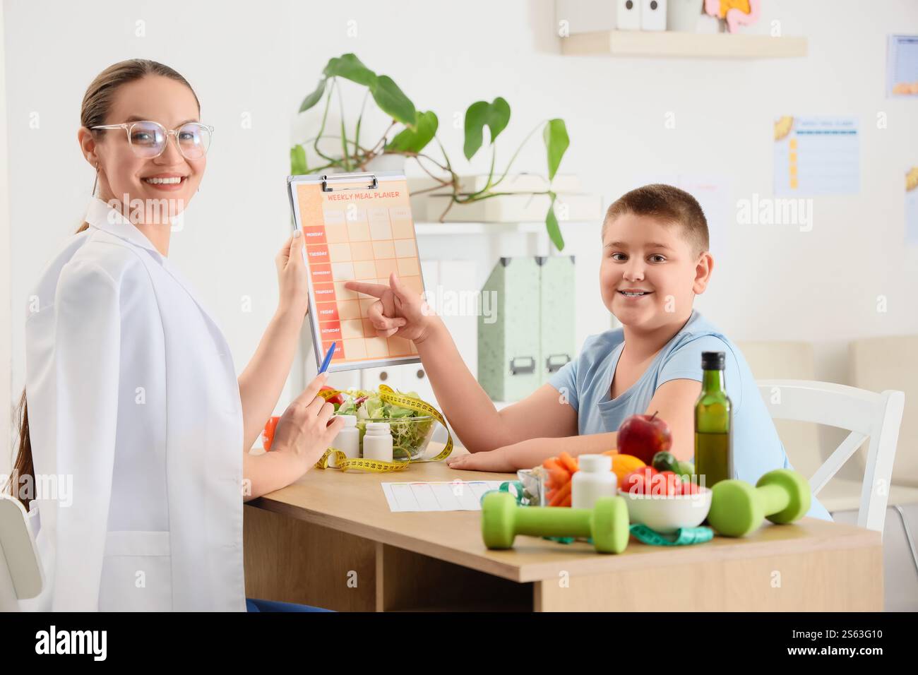 Overweight boy and nutritionist with meal planner at table in clinic ...