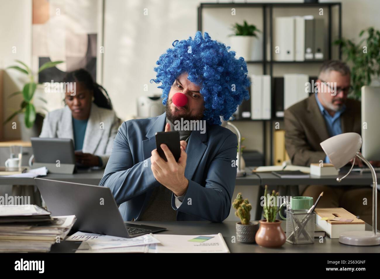Young clown employee wagging finger to colleague on smartphone screen ...