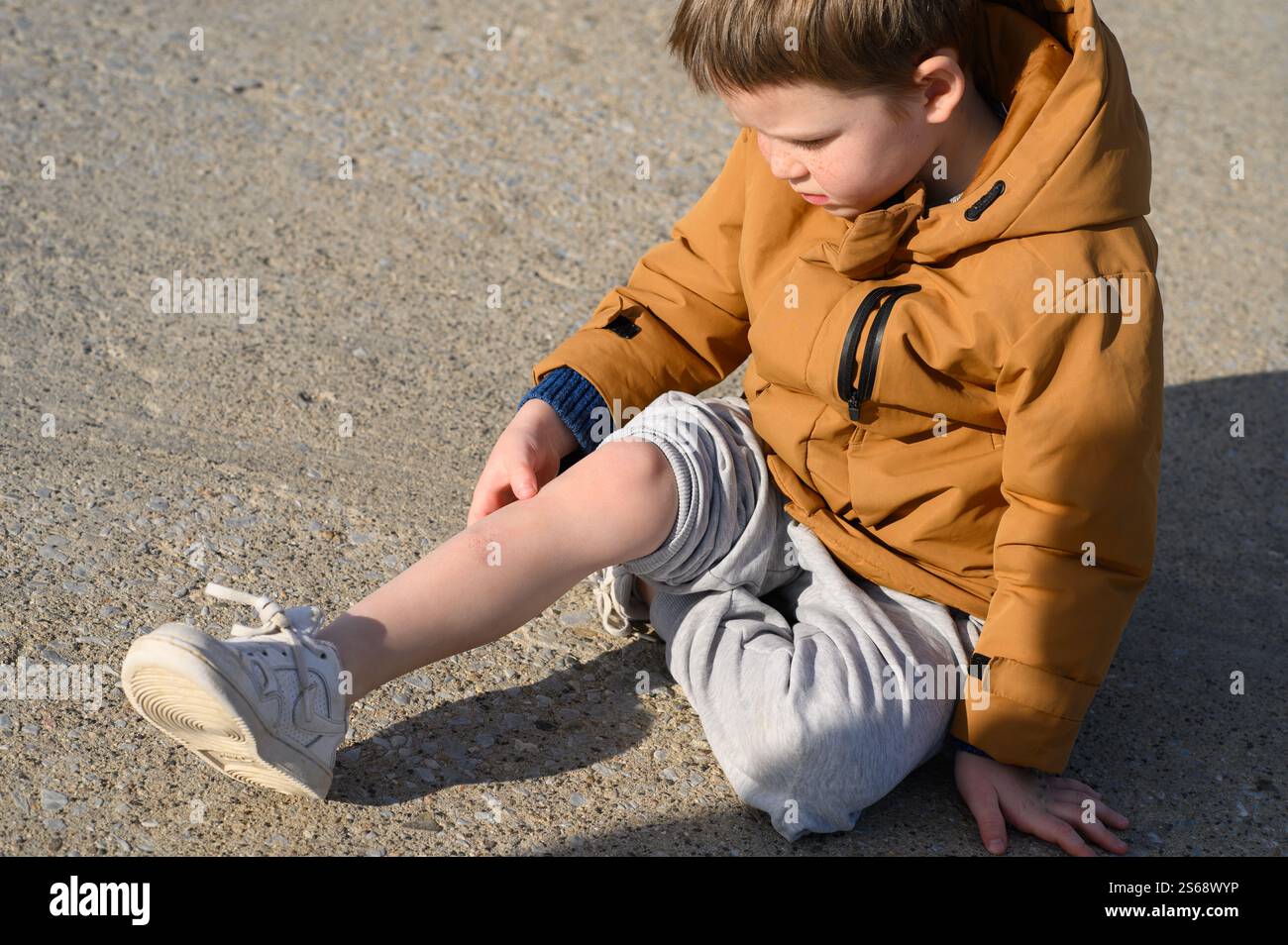 A child sits on the asphalt with a scraped knee, looking upset after a ...