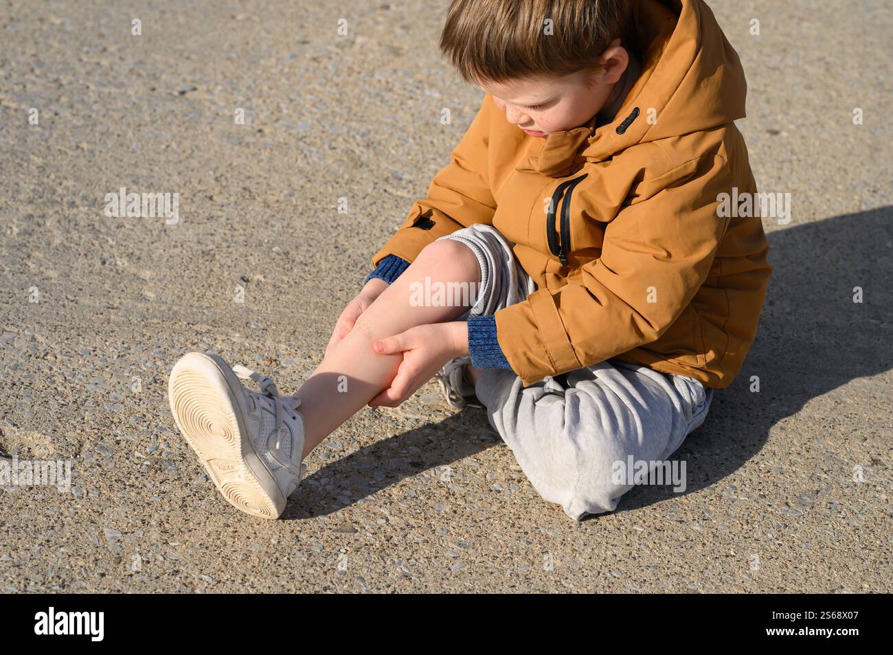 A child sits on the asphalt with a scraped knee, looking upset after a ...
