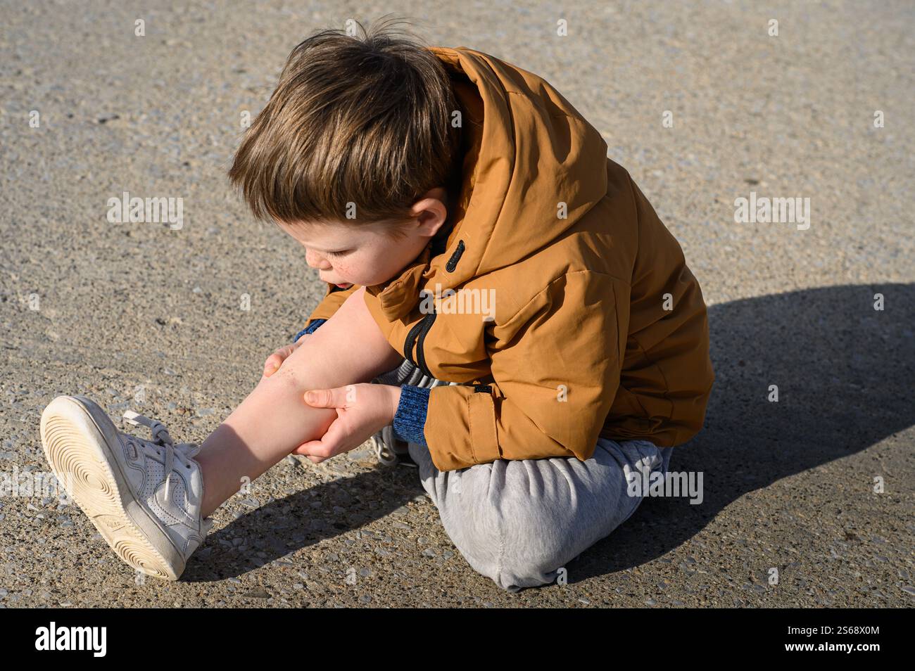 A child sits on the asphalt with a scraped knee, looking upset after a ...