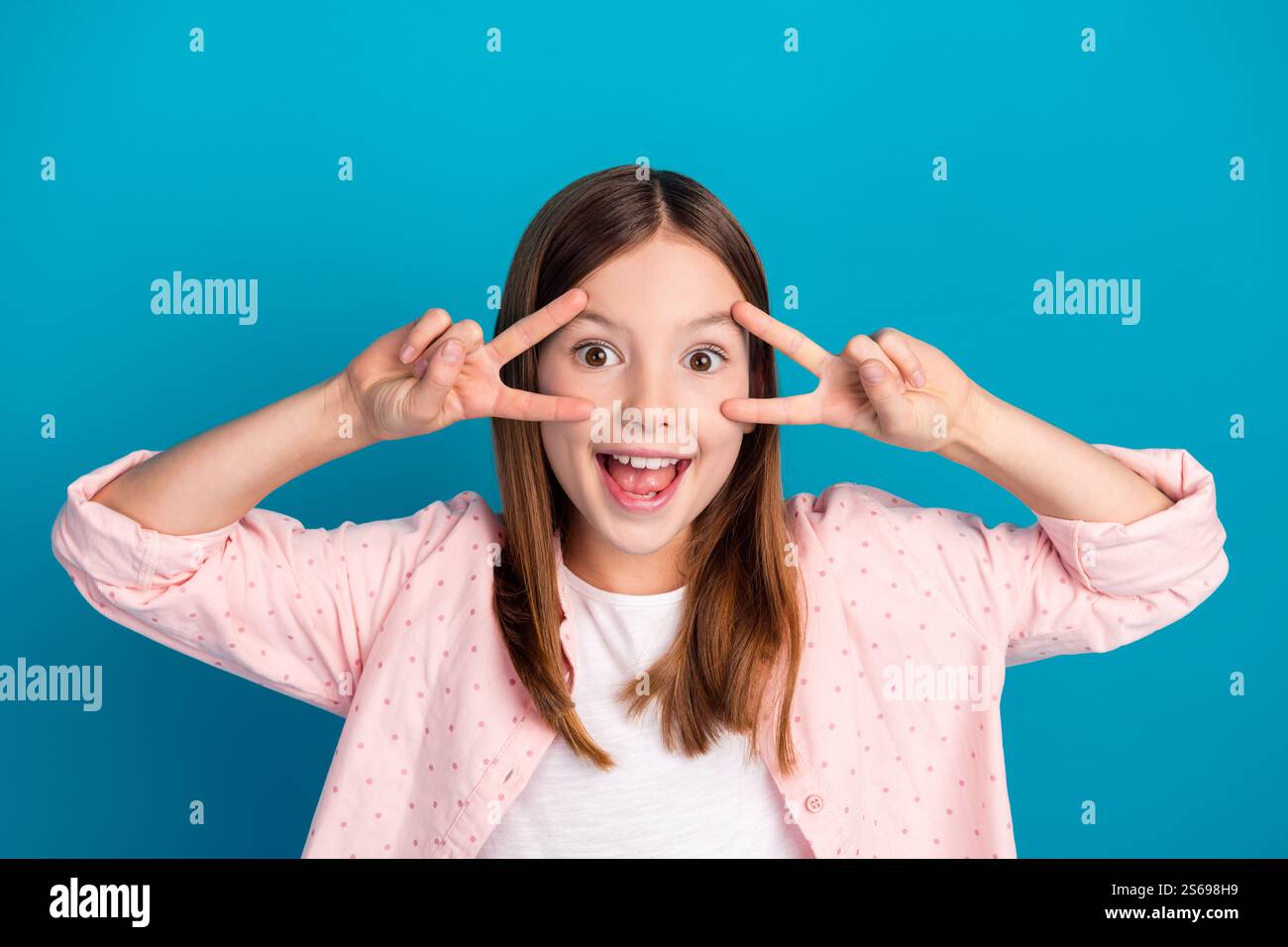 Smiling young girl making a playful hand sign against a bright blue ...