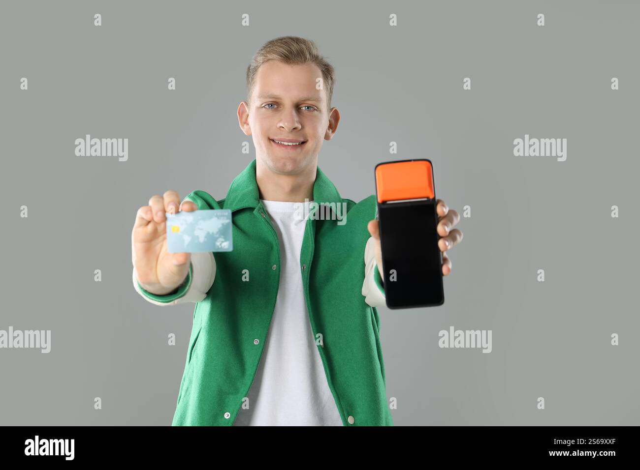 Happy young man with payment terminal and debit card on light grey ...