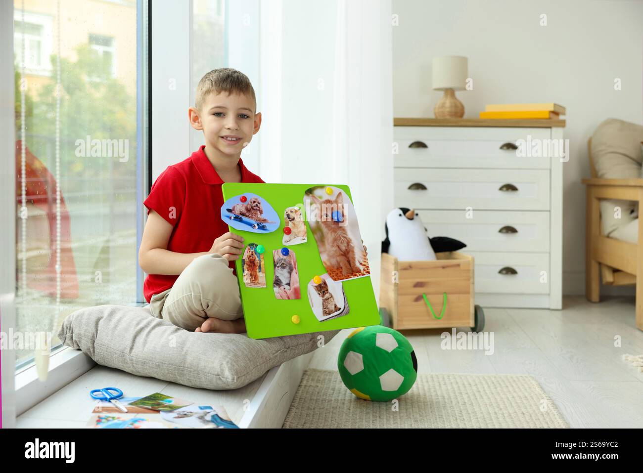 Little boy holding vision board with different pictures near window ...