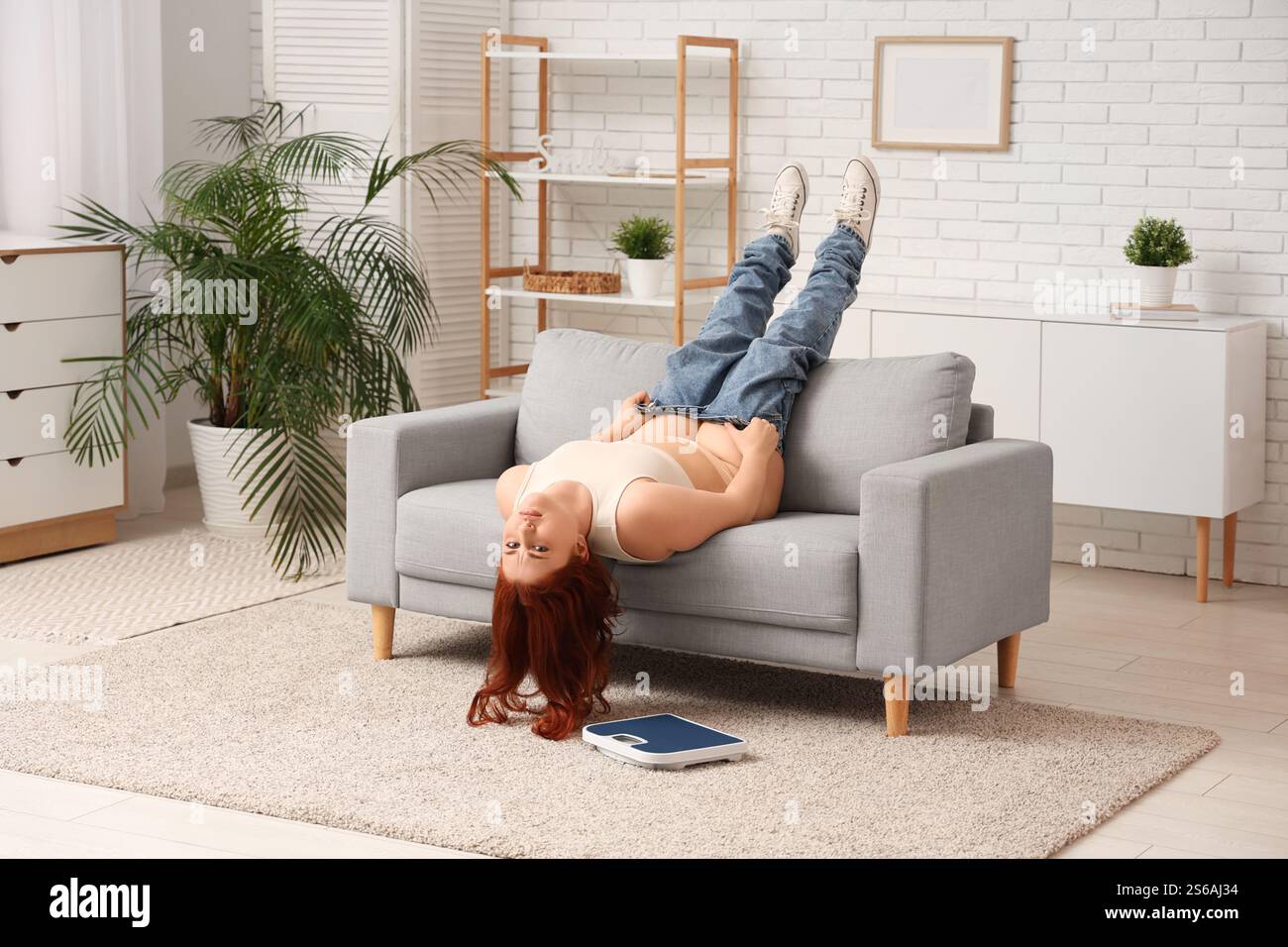 Young woman trying to put tight jeans on sofa at home. Weight gain ...
