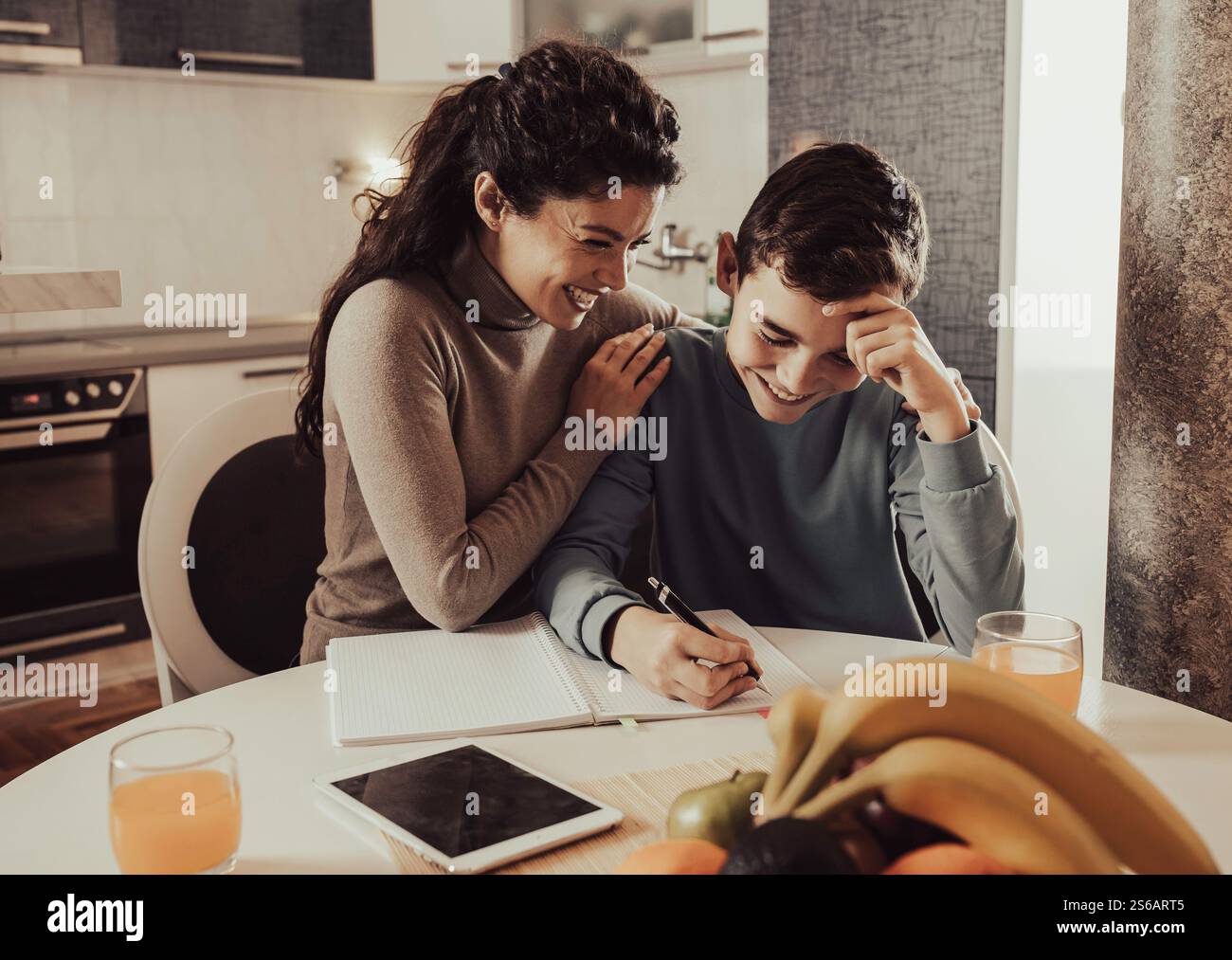 Mother sitting with son while doing homework at dining table in kitchen ...