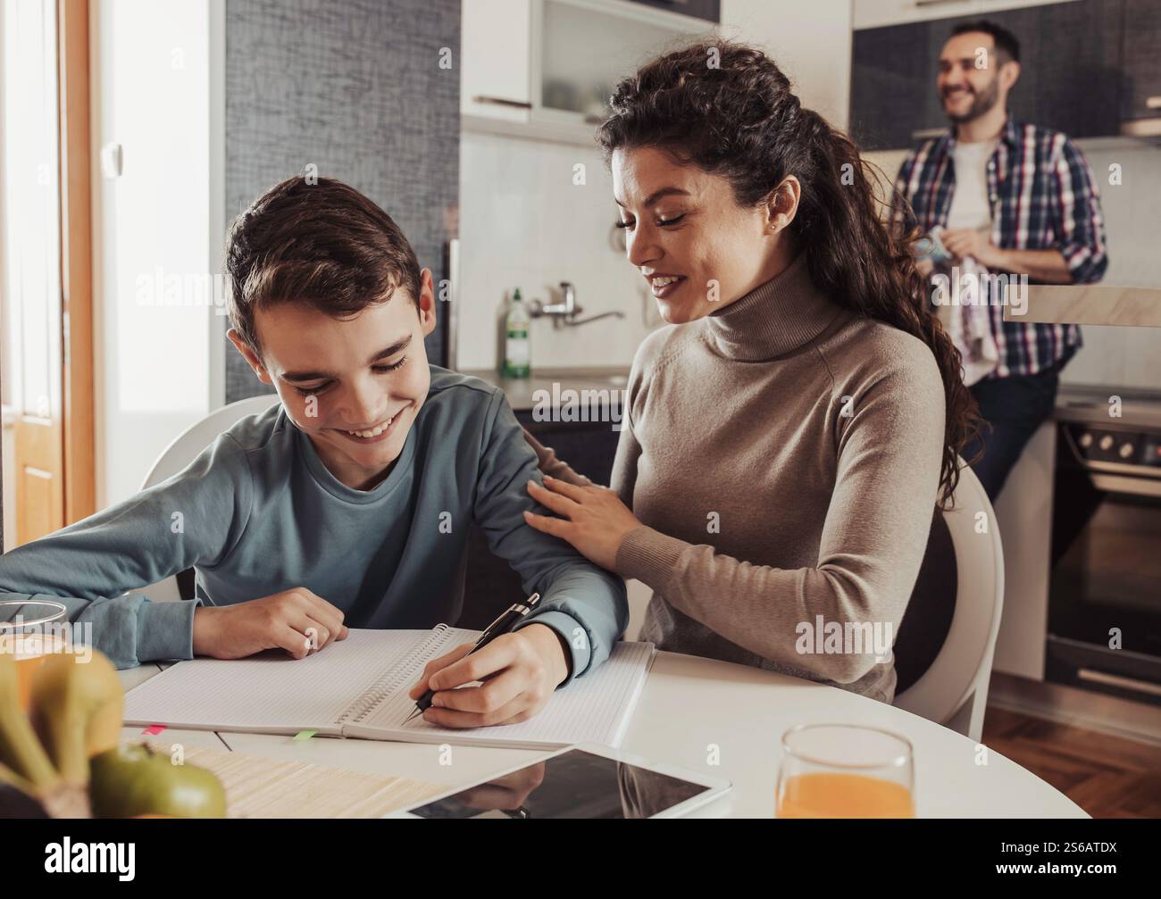 Mother sitting with son while doing homework on table in kitchen, smiling, happy, satisfied ...