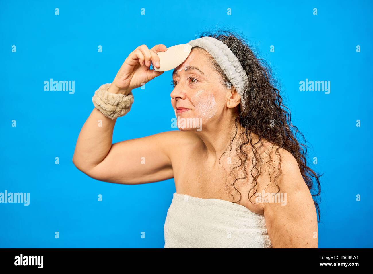 Mature woman using a sponge to apply face cream, promoting skincare for ...