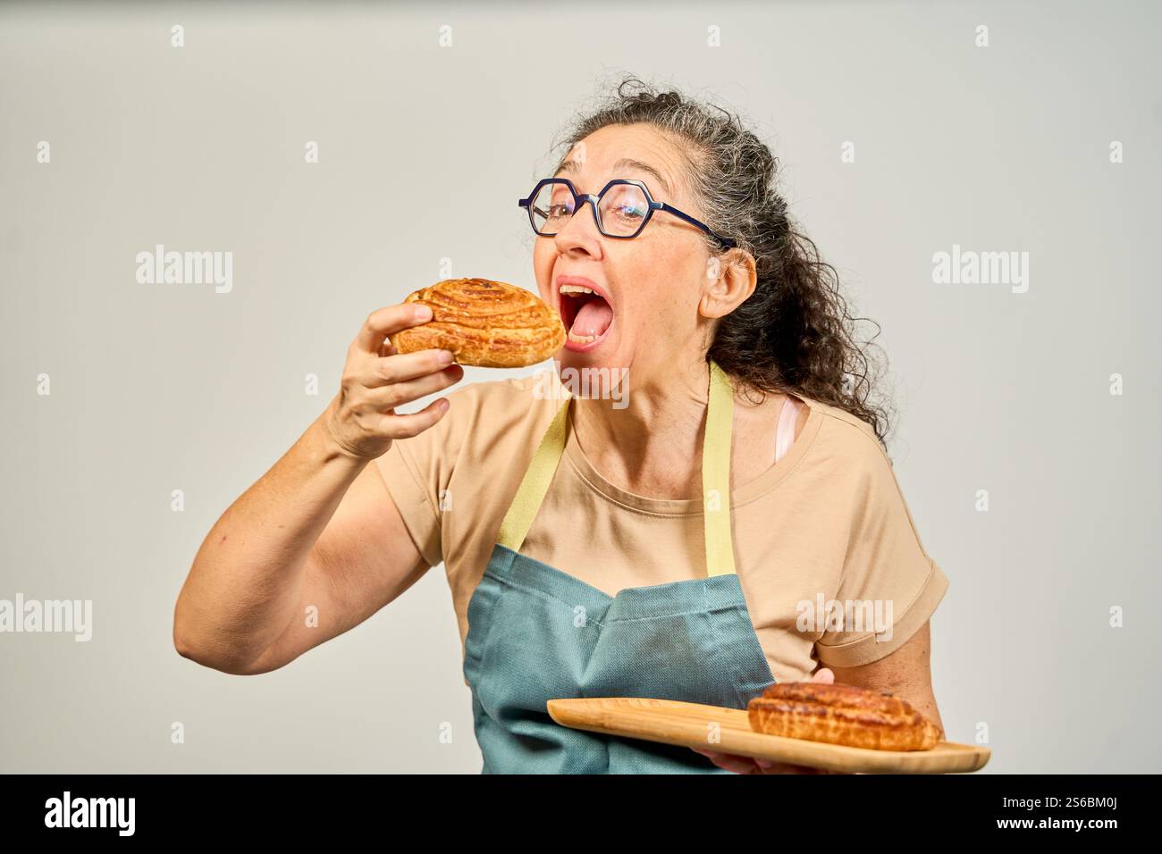 A woman is eating a donut and is wearing an apron. She is smiling and ...