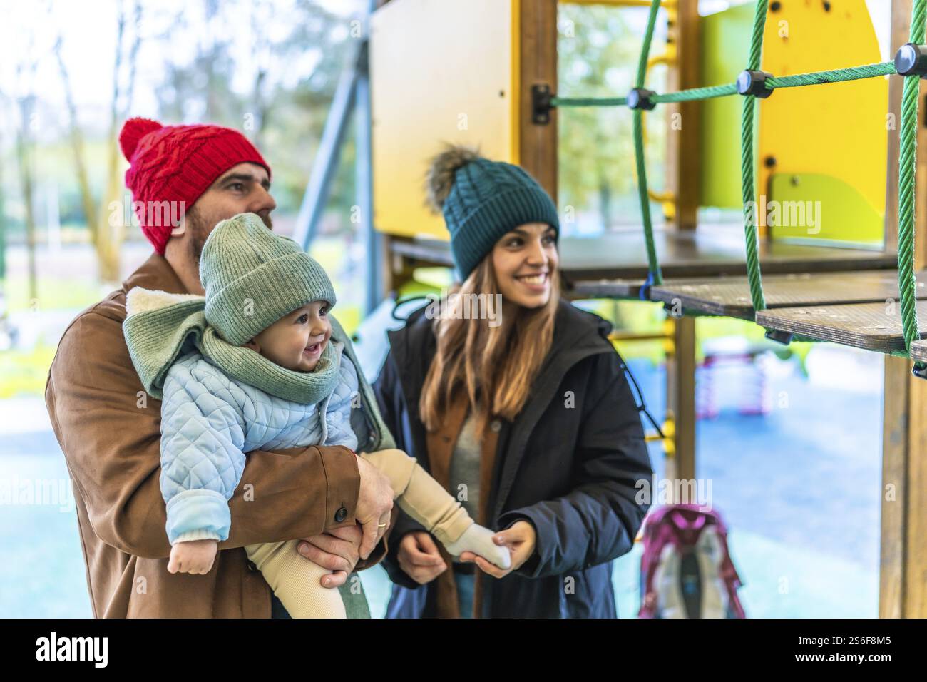 Smiling parents holding their baby at the playground, wearing winter ...