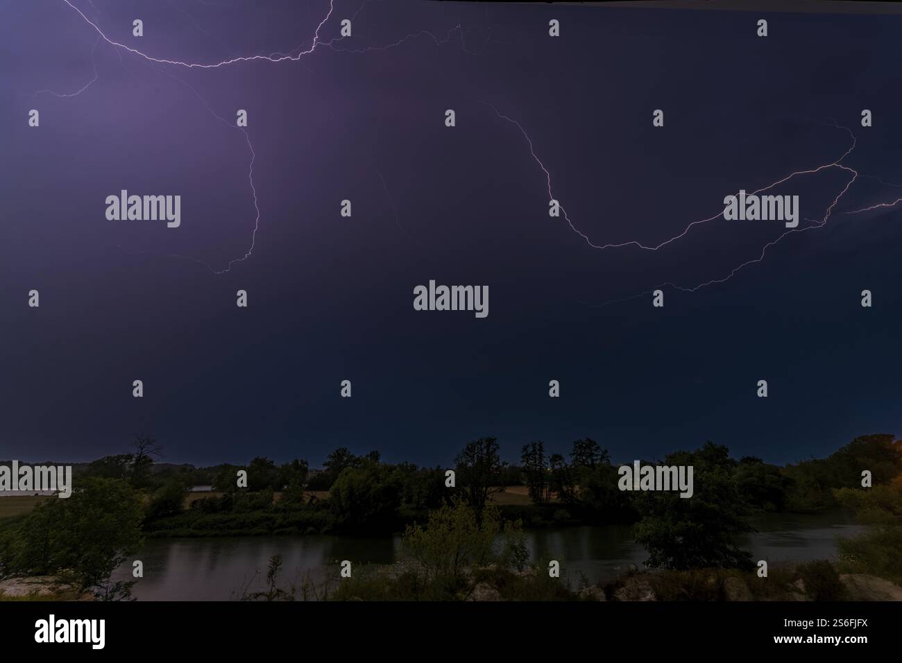 Lightning illuminates the dark night scene above a wooded riverbank ...