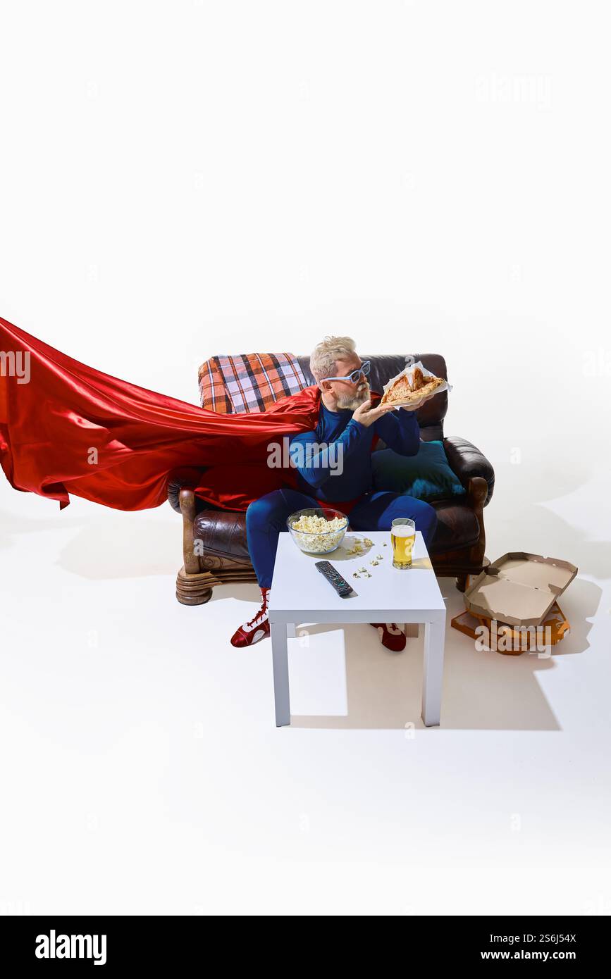 Grey-haired man in superhero costume sitting on couch, eating pizza ...