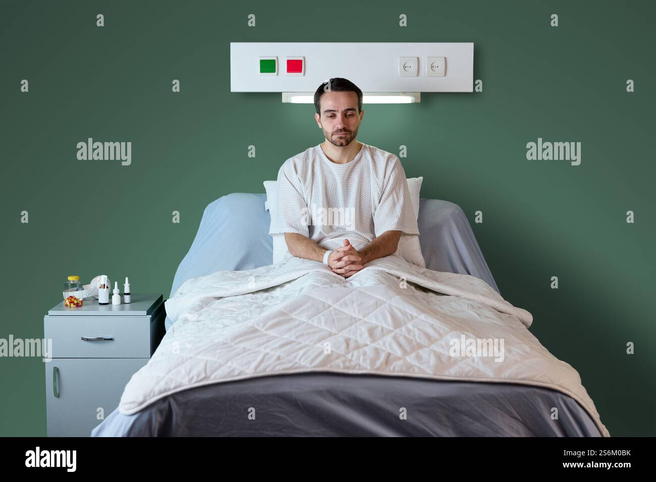 Young man, patient sitting in bed with blanket in hospital gown with ...