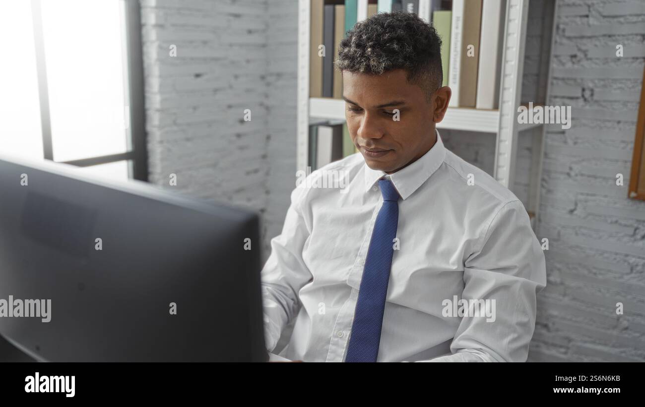Young hispanic man working on a computer in an office setting, dressed in formal attire, focused ...