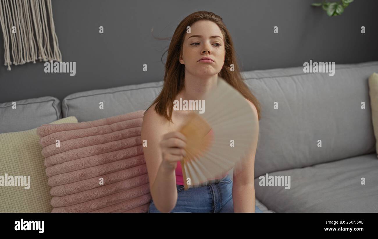 Attractive young redhead woman fanning herself while sitting in a cozy ...