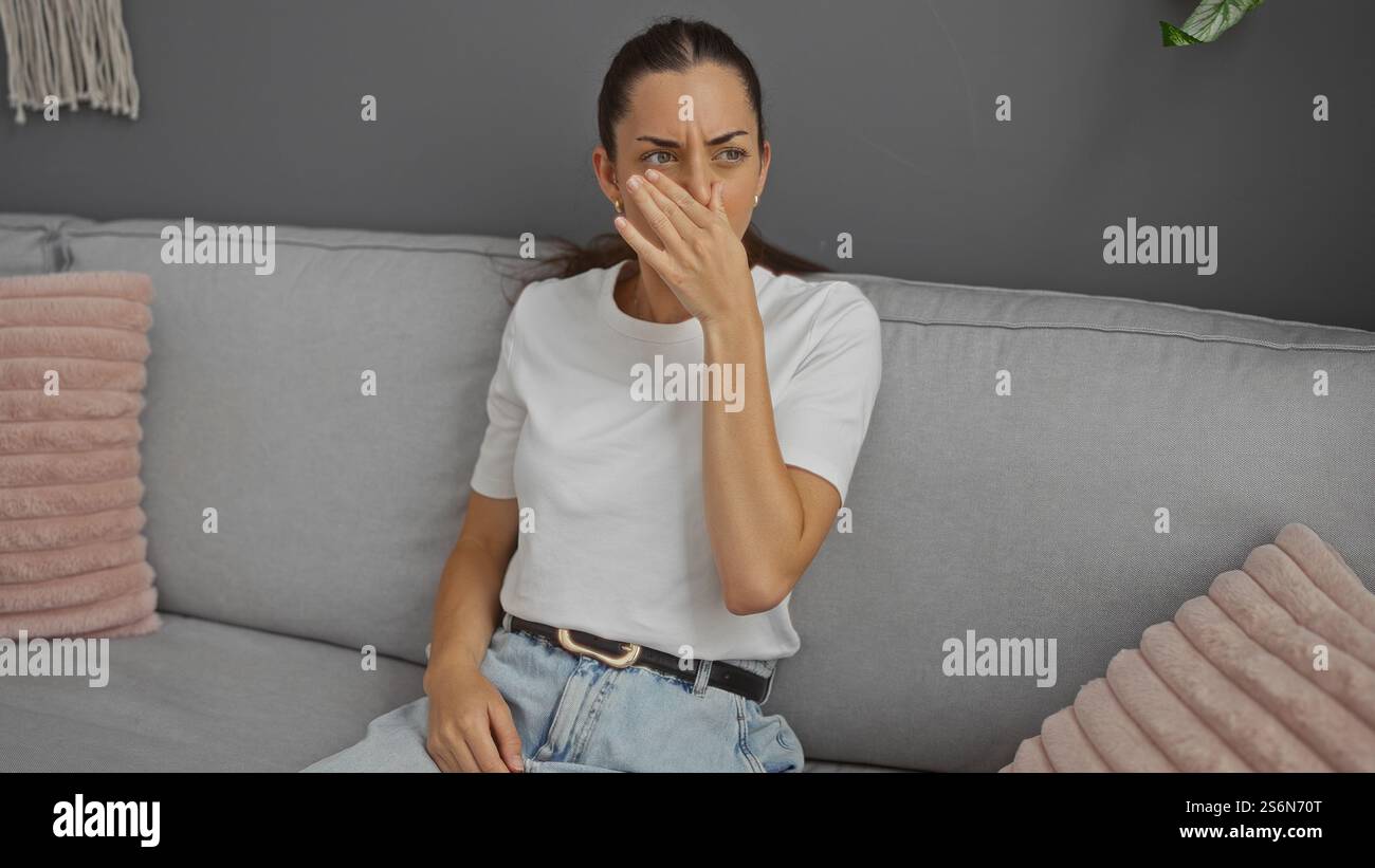 Woman sitting on a couch in a neutral-toned living room, covering her nose with her hand while ...