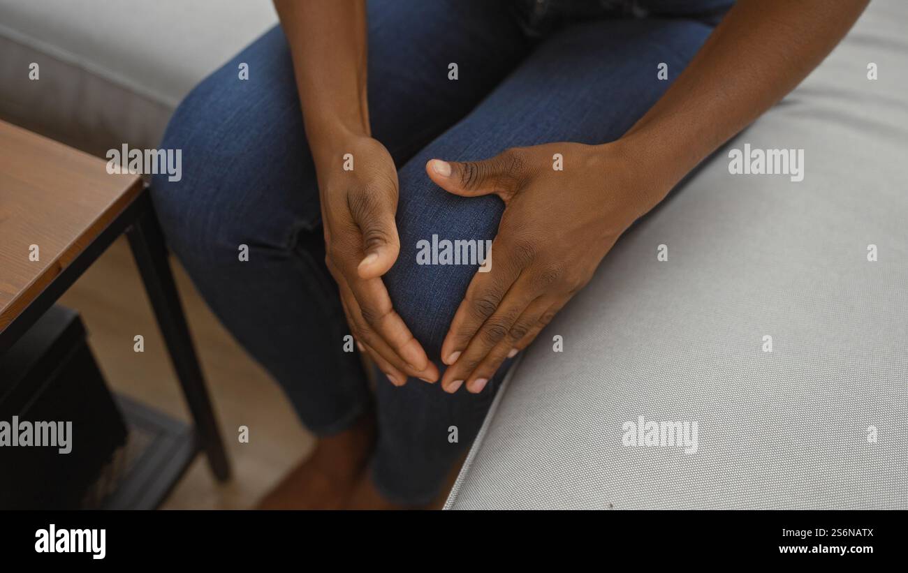 Young woman with knee pain sitting in living room, wearing jeans, hands ...