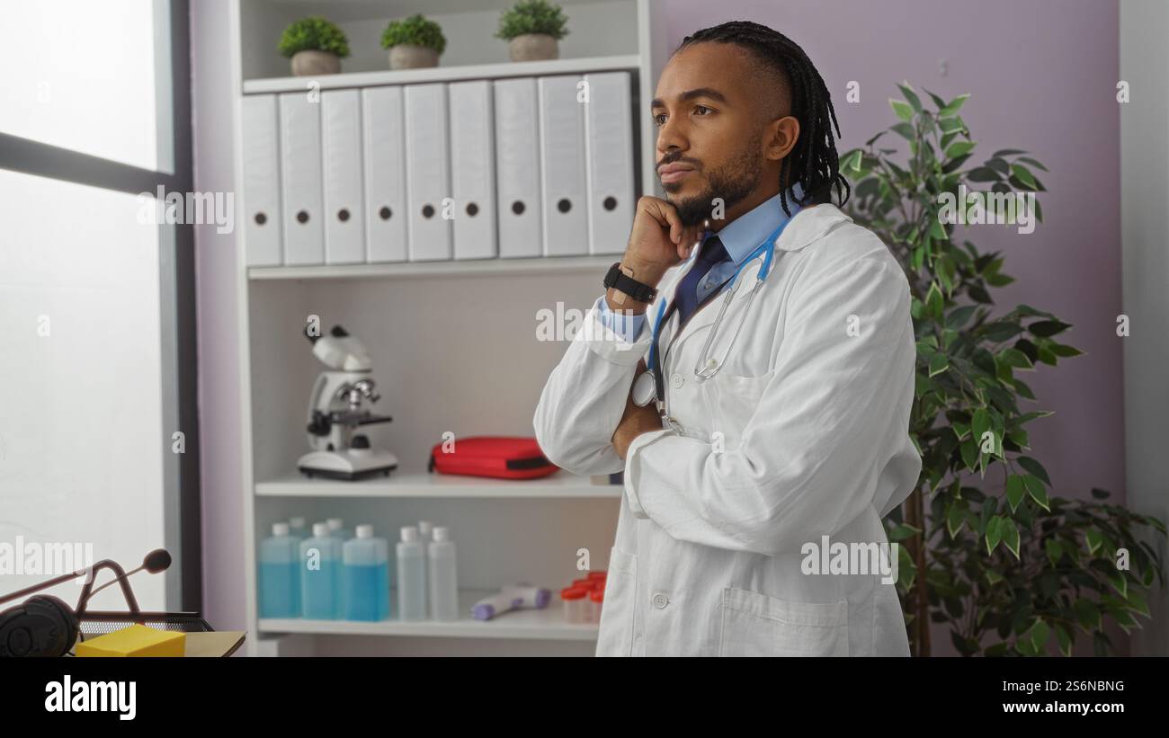 Young man with braids wearing lab coat in clinic standing thoughtfully ...