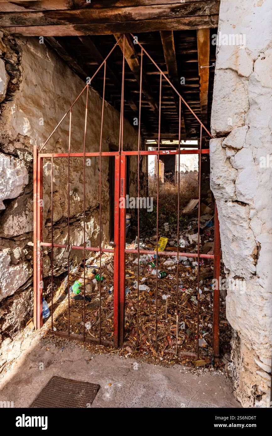 A red gate is open in a dirty alleyway. The gate is rusted and has a ...
