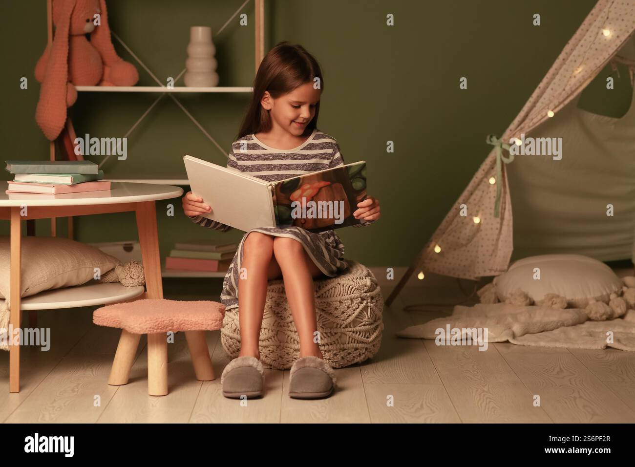 Cute little girl reading fairy-tale while sitting on pouf at home Stock ...